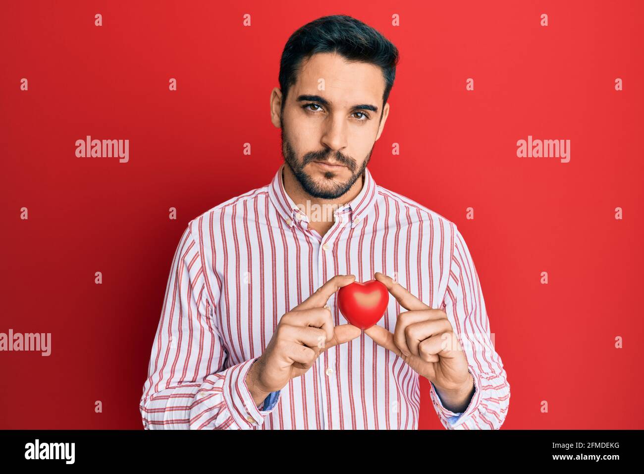 Young hispanic man holding heart relaxed with serious expression on ...