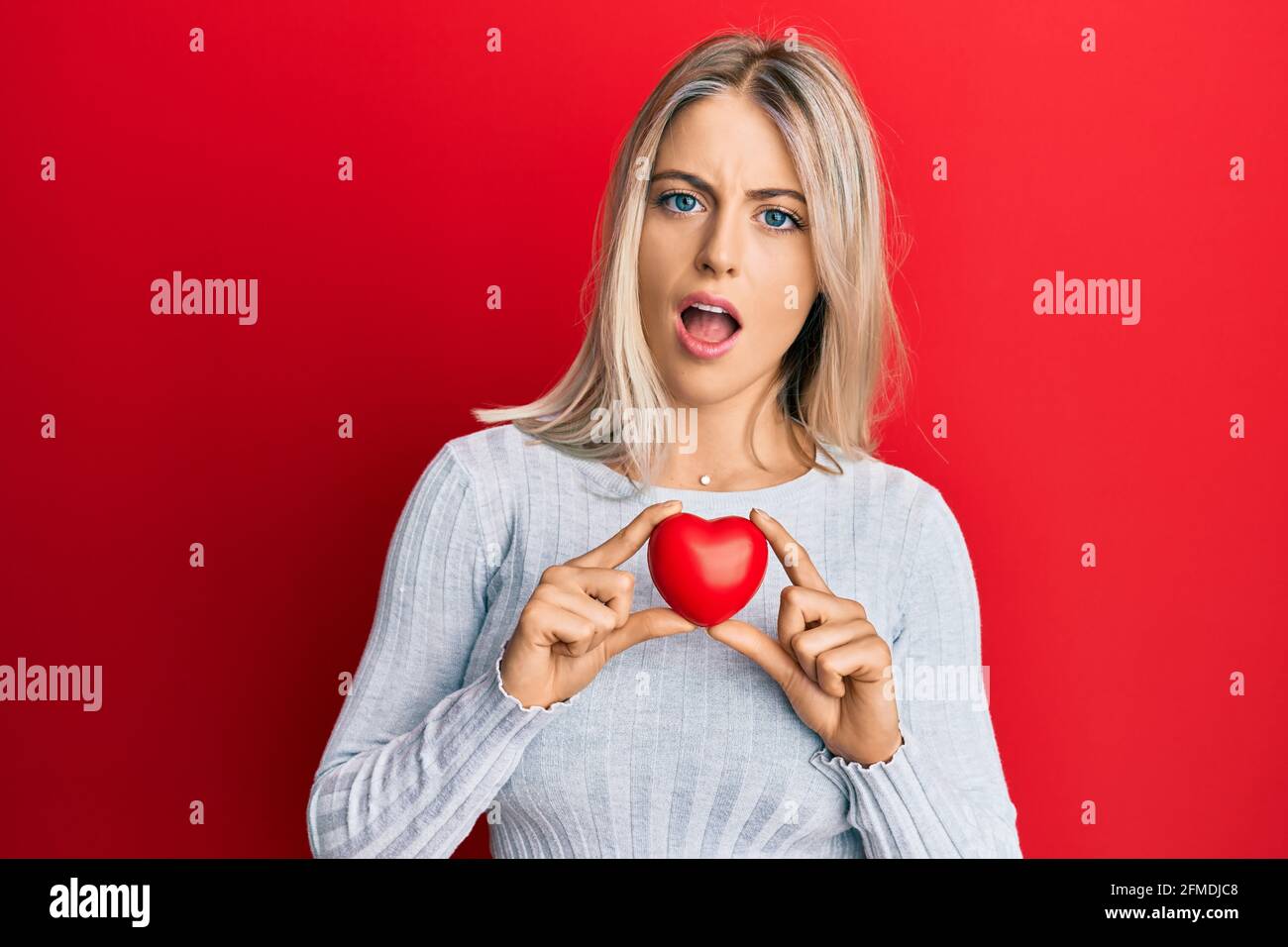 Beautiful blonde woman holding heart in shock face, looking skeptical ...