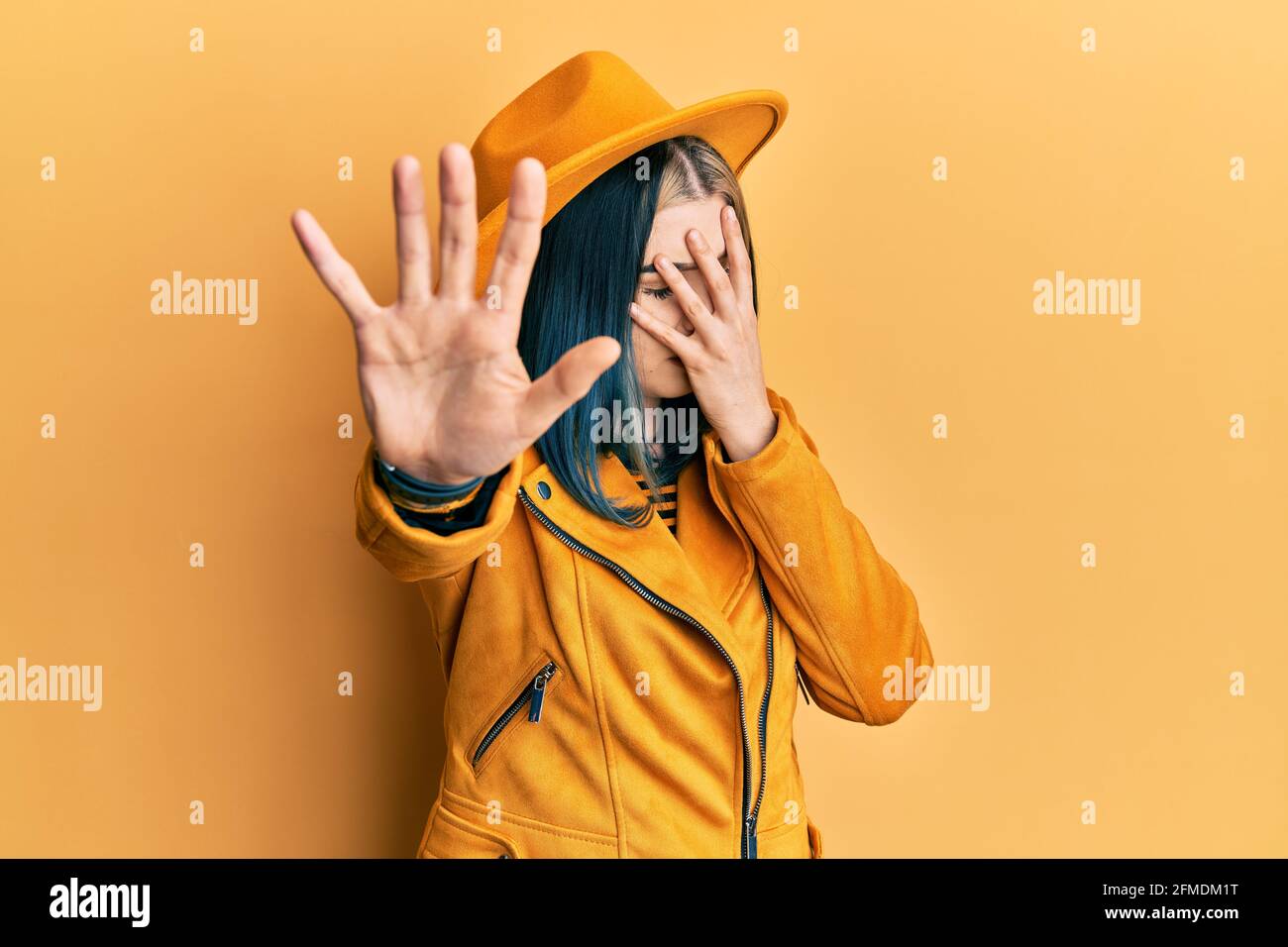 Young modern girl wearing yellow hat and leather jacket covering eyes ...