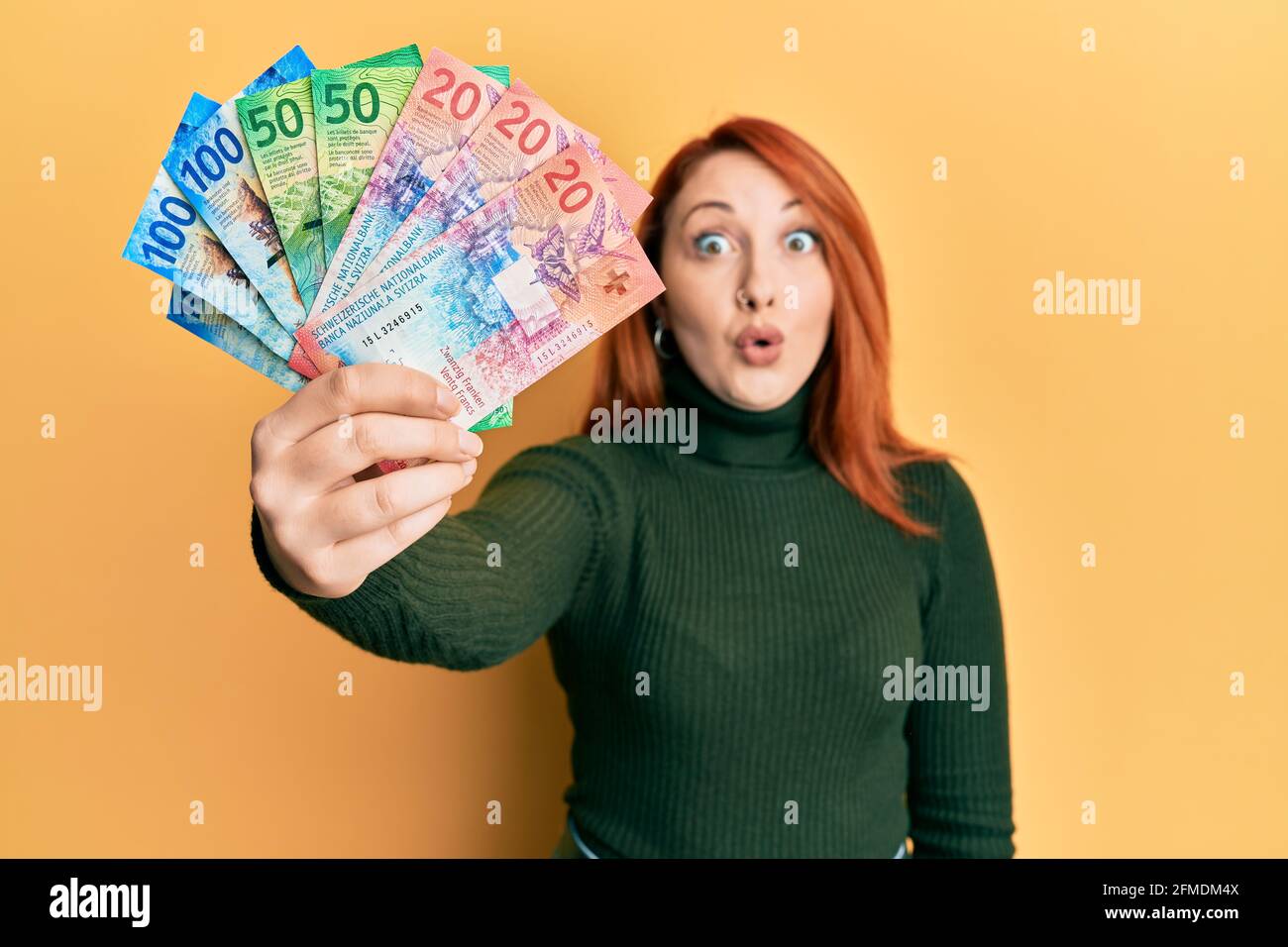 Beautiful redhead woman holding swiss franc banknotes scared and amazed ...