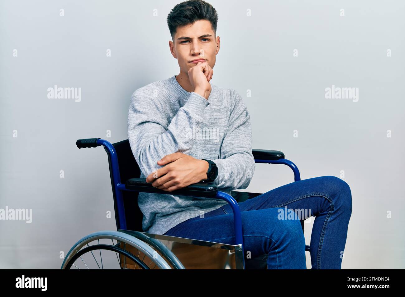 Young hispanic man sitting on wheelchair with hand on chin thinking ...