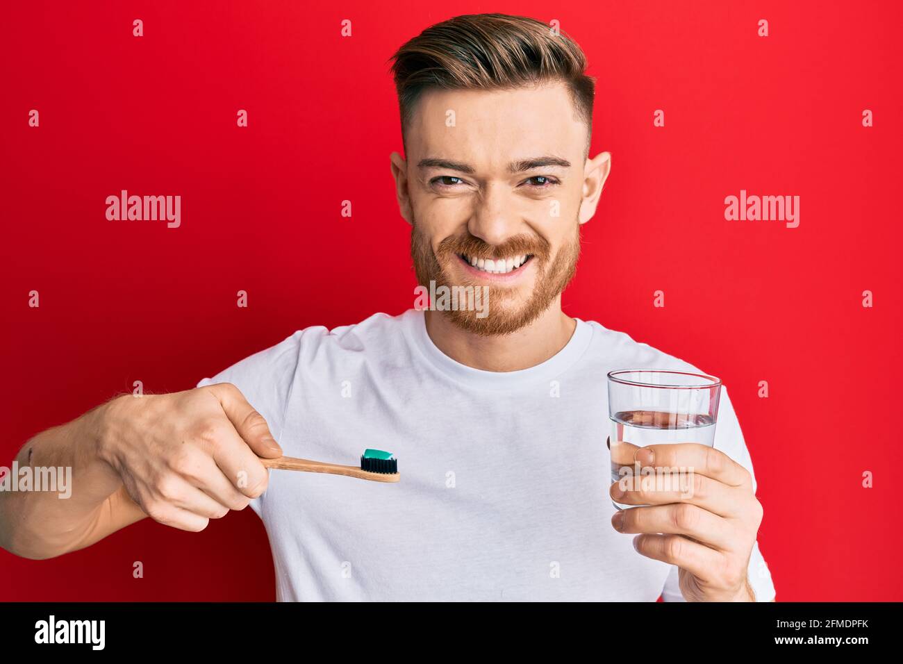 Young redhead man holding toothbrush with toothpaste and glass of water ...