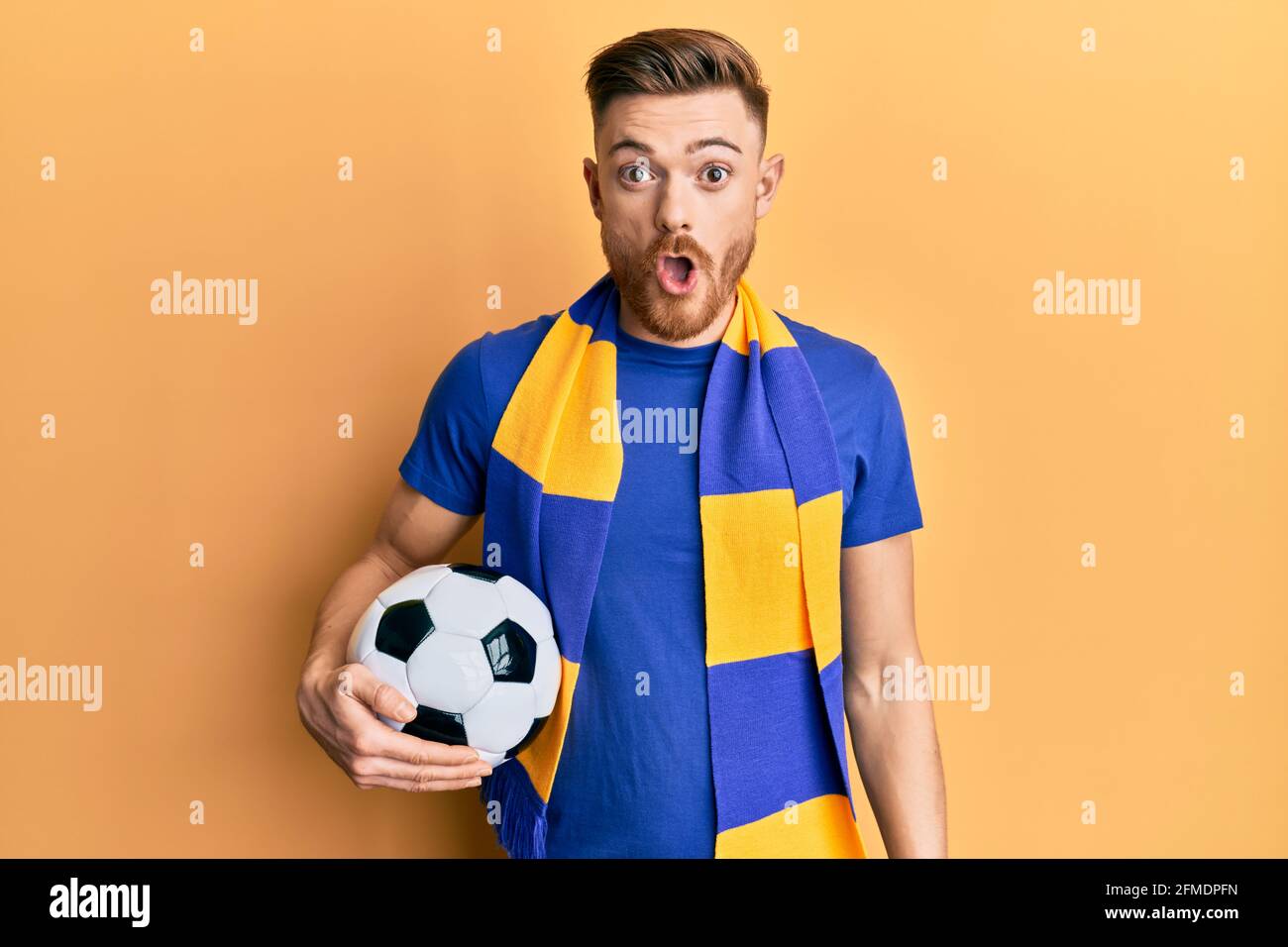 Young redhead man football hooligan holding ball scared and amazed with ...