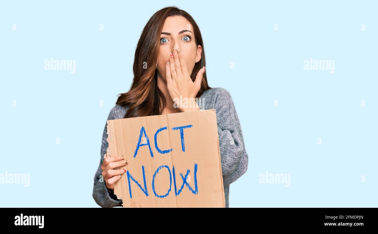 Young brunette woman holding act now banner covering mouth with hand ...