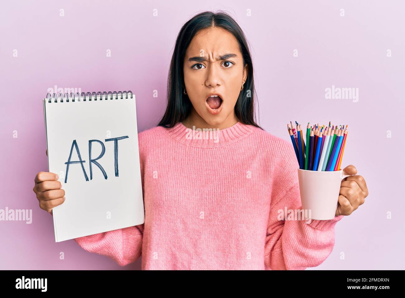 Young asian woman holding art notebook and colored pencils in shock ...