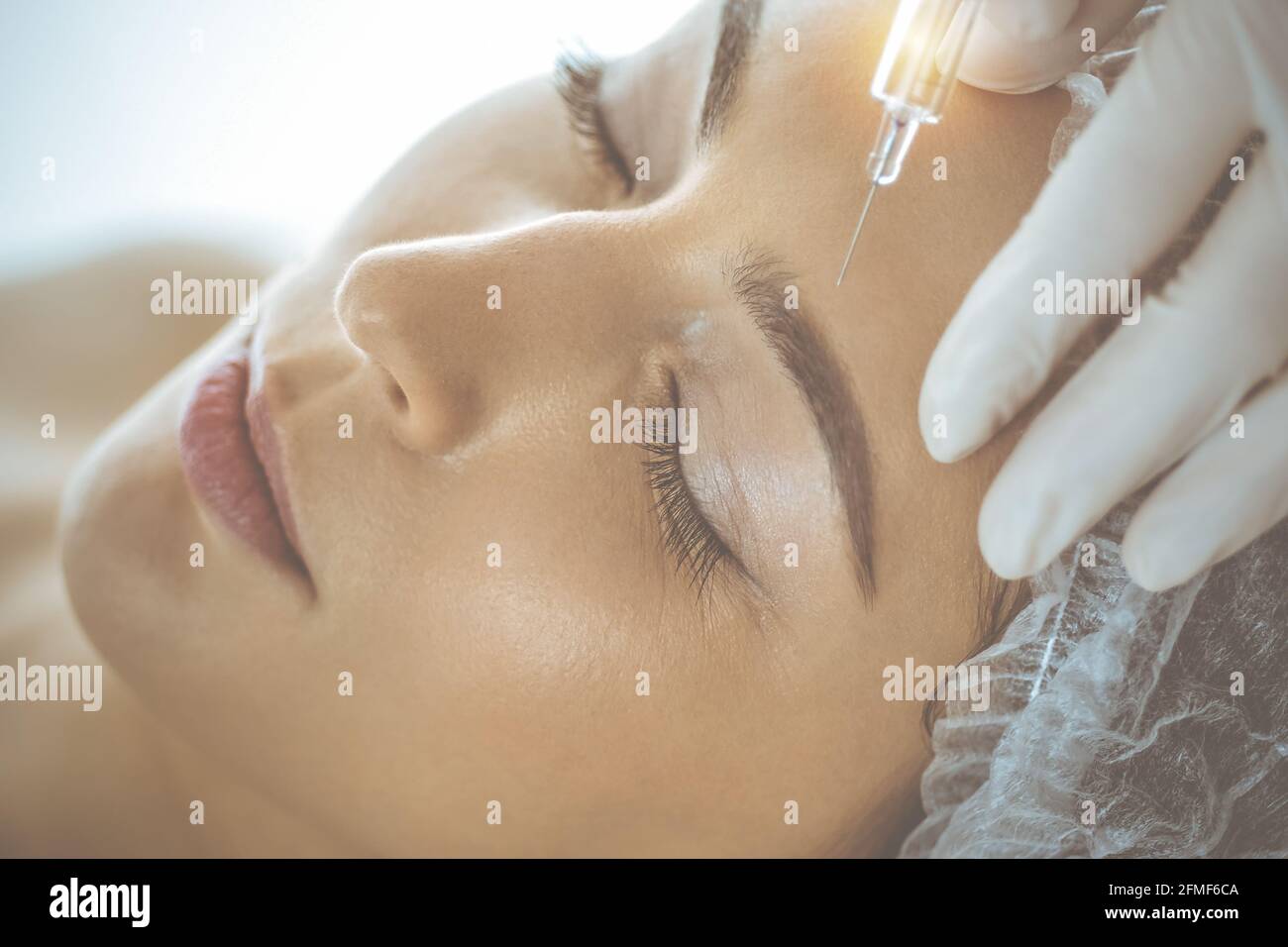Beautician doing beauty procedure with syringe to face of young brunette woman in sunny clinic ...