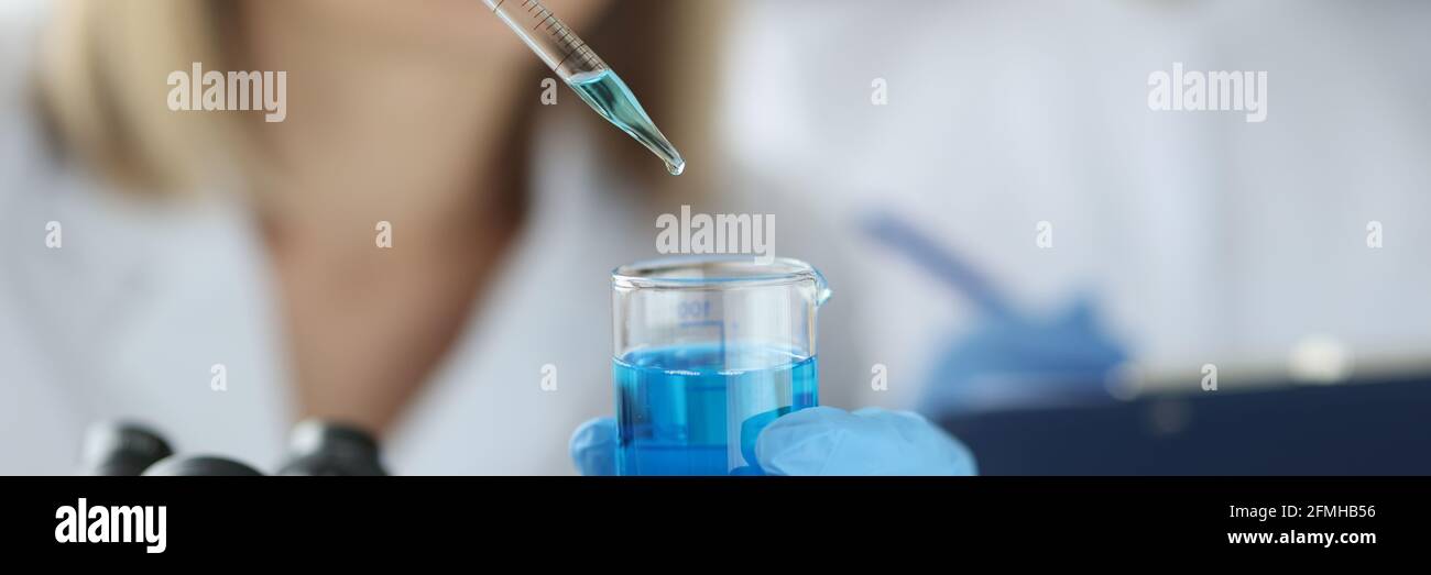 Female scientist dripping blue liquid from pipette into flask in ...
