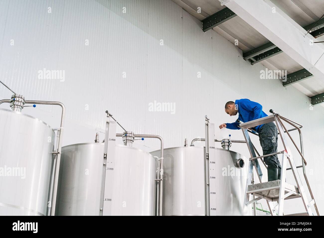 Male engineer on ladder pouring liquid into stainless steel tank while ...