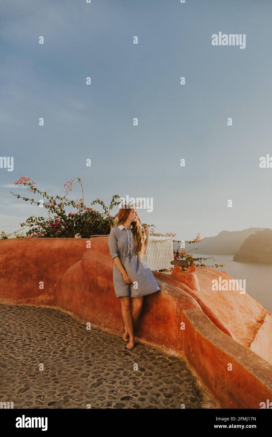 Full length young female in sundress sitting on coastal town embankment ...