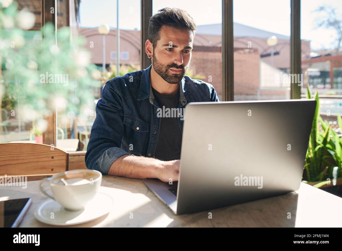 Bearded ethnic male remote employee typing on netbook at table with cup ...