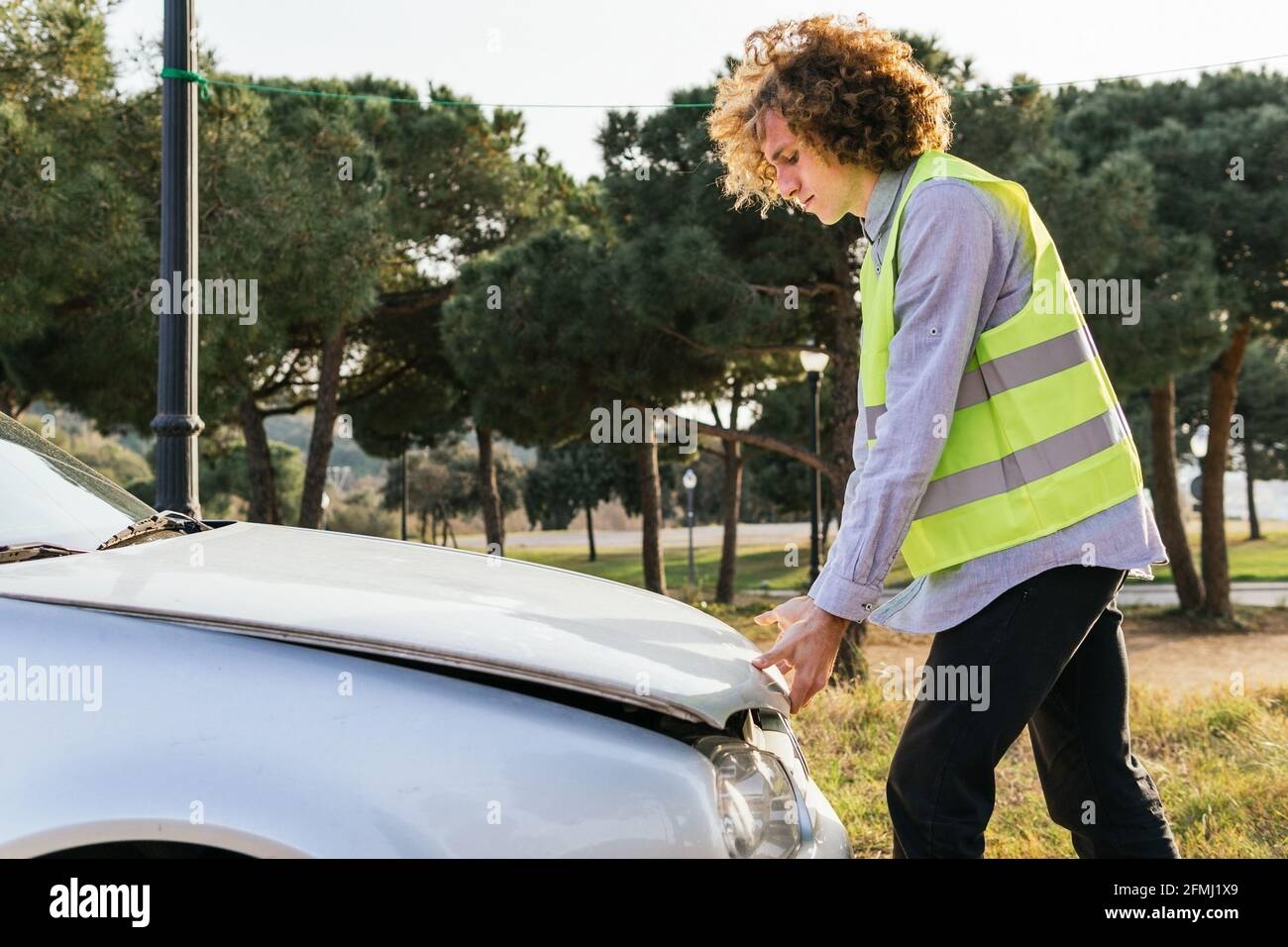 Side view of young male driver in safety vest opening car hood while ...