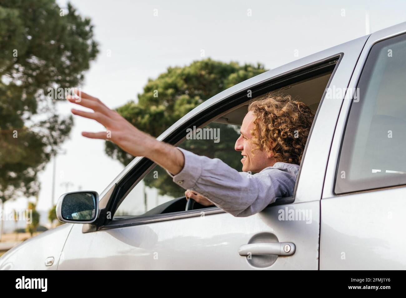Side view of cheerful young haired male keeping hand out of car window ...