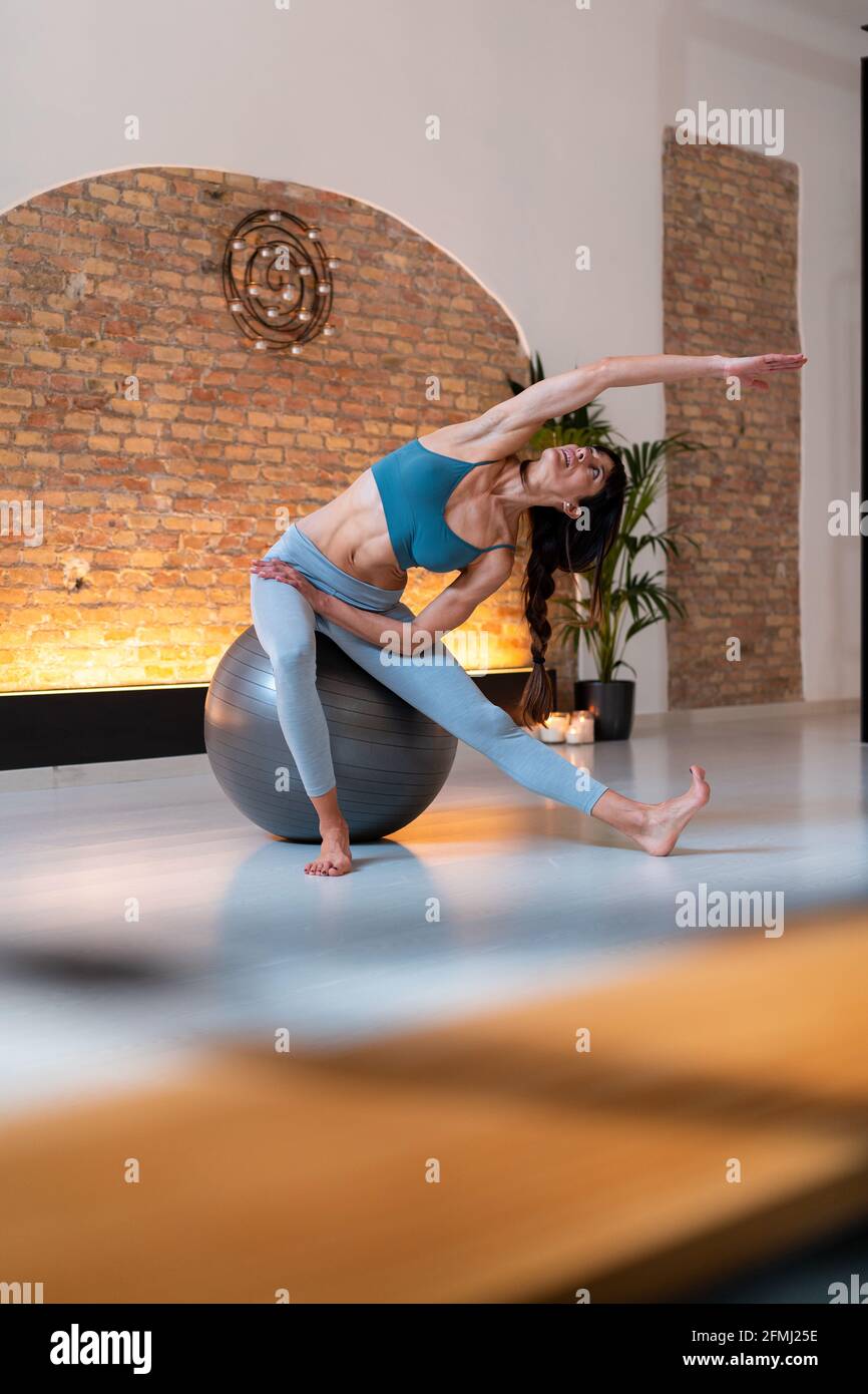 Flexible athletic female sitting on fitness ball and doing side bend ...