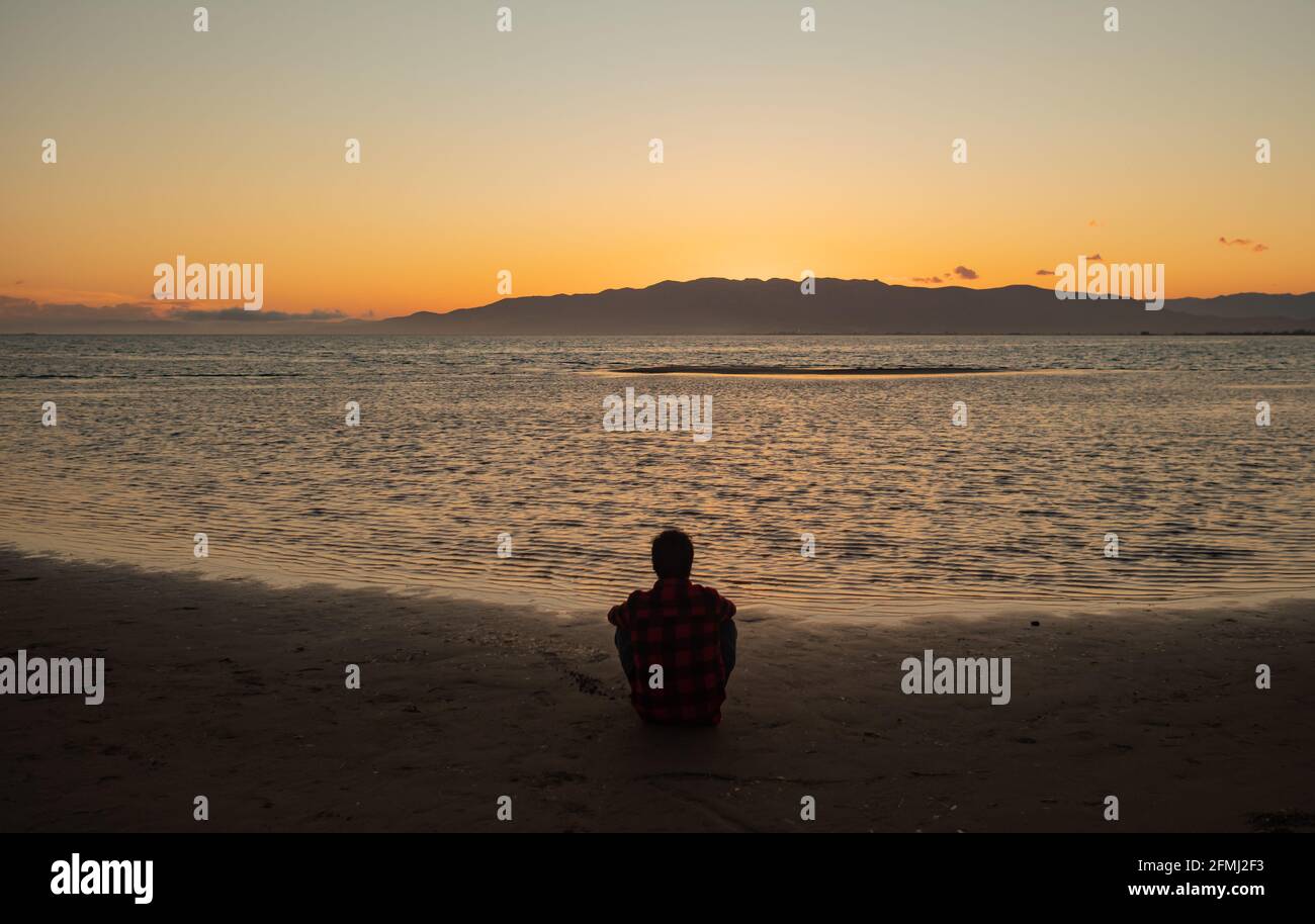 Back view of unrecognizable tourist sitting alone on sandy beach near ...