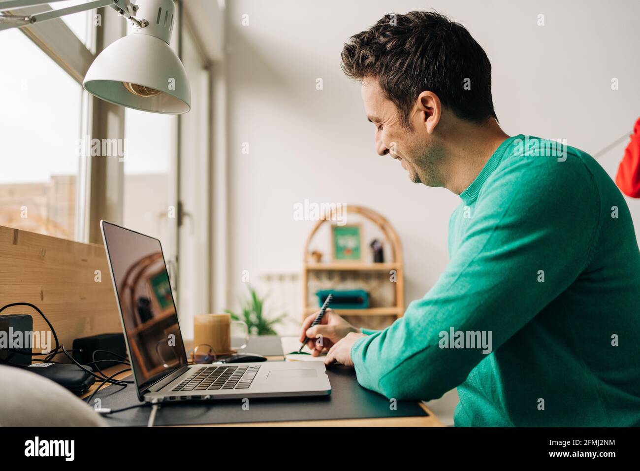 Side view of cheerful male remote worker taking notes against netbook ...
