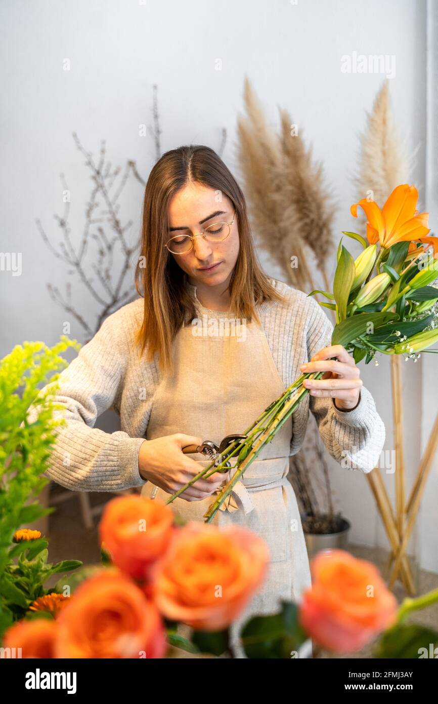 Focused young ethnic female florist in casual clothes and apron cutting ...