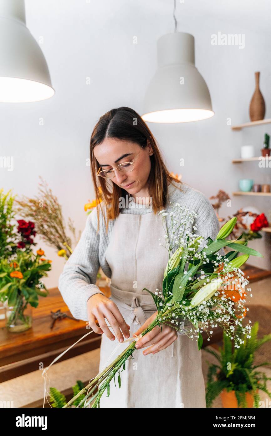 Focused young ethnic female florist in casual clothes and apron tying ...