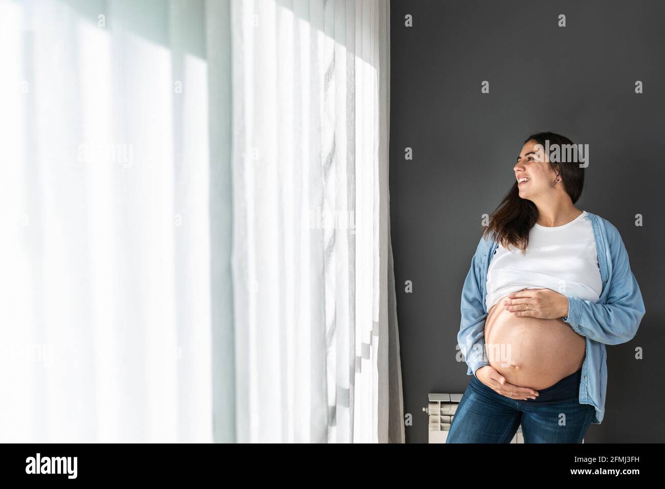 Positive pregnant female gently touching tummy while standing in room ...