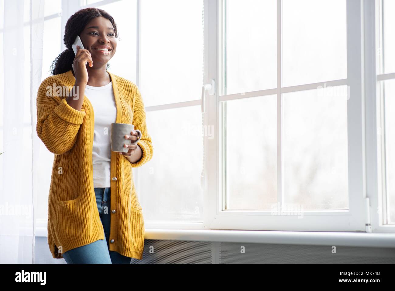 Pretty young black lady with mug standing by window and talking on ...