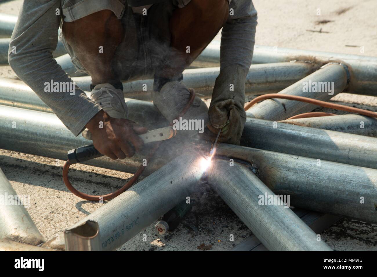 A welder is sitting on a steel roof frame and is welding steel pipes ...