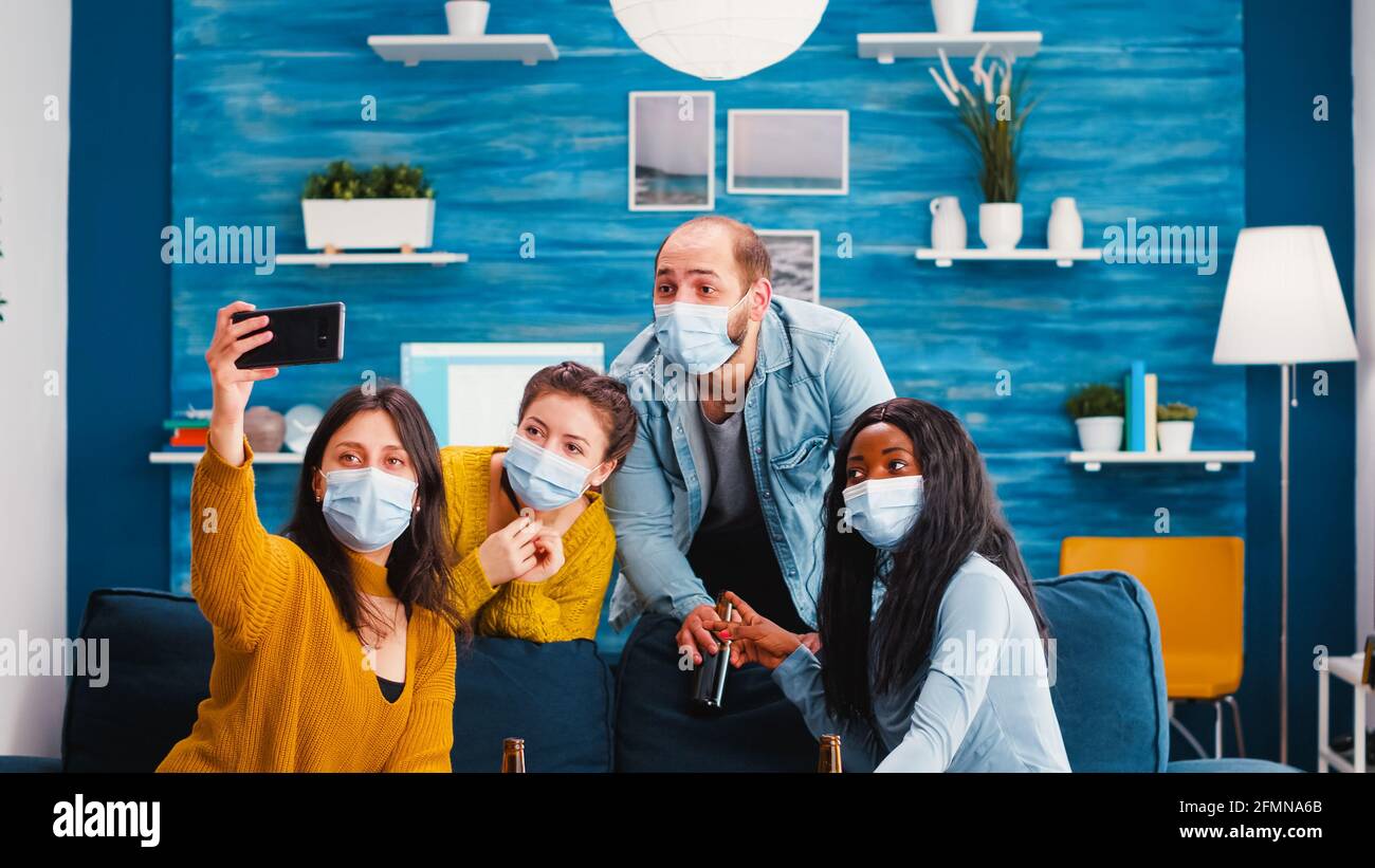 Smiling multiracial friends sitting on sofa with protection masks ...