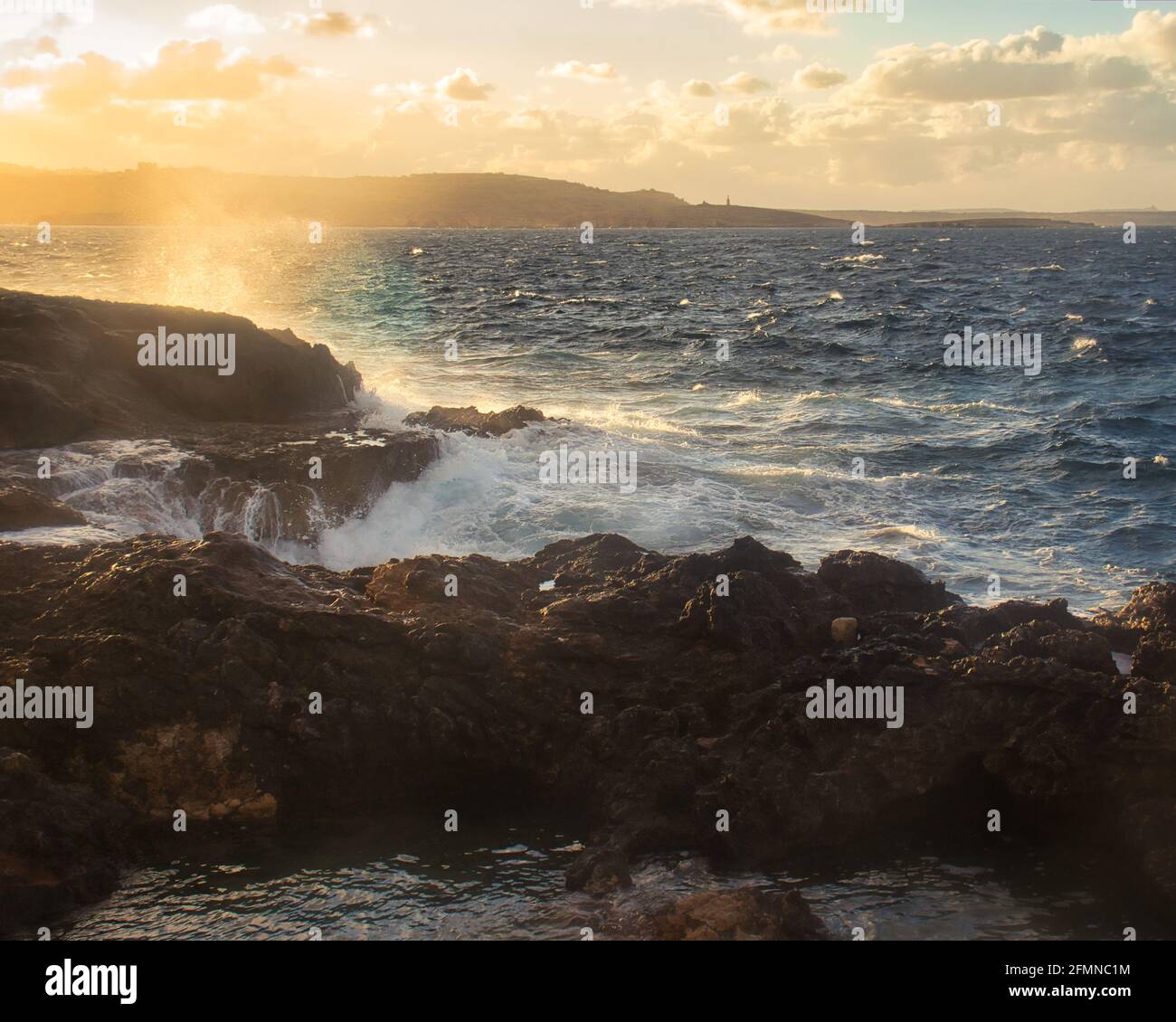 Golden light hitting water spraying over rocks on a beach in Qawra ...