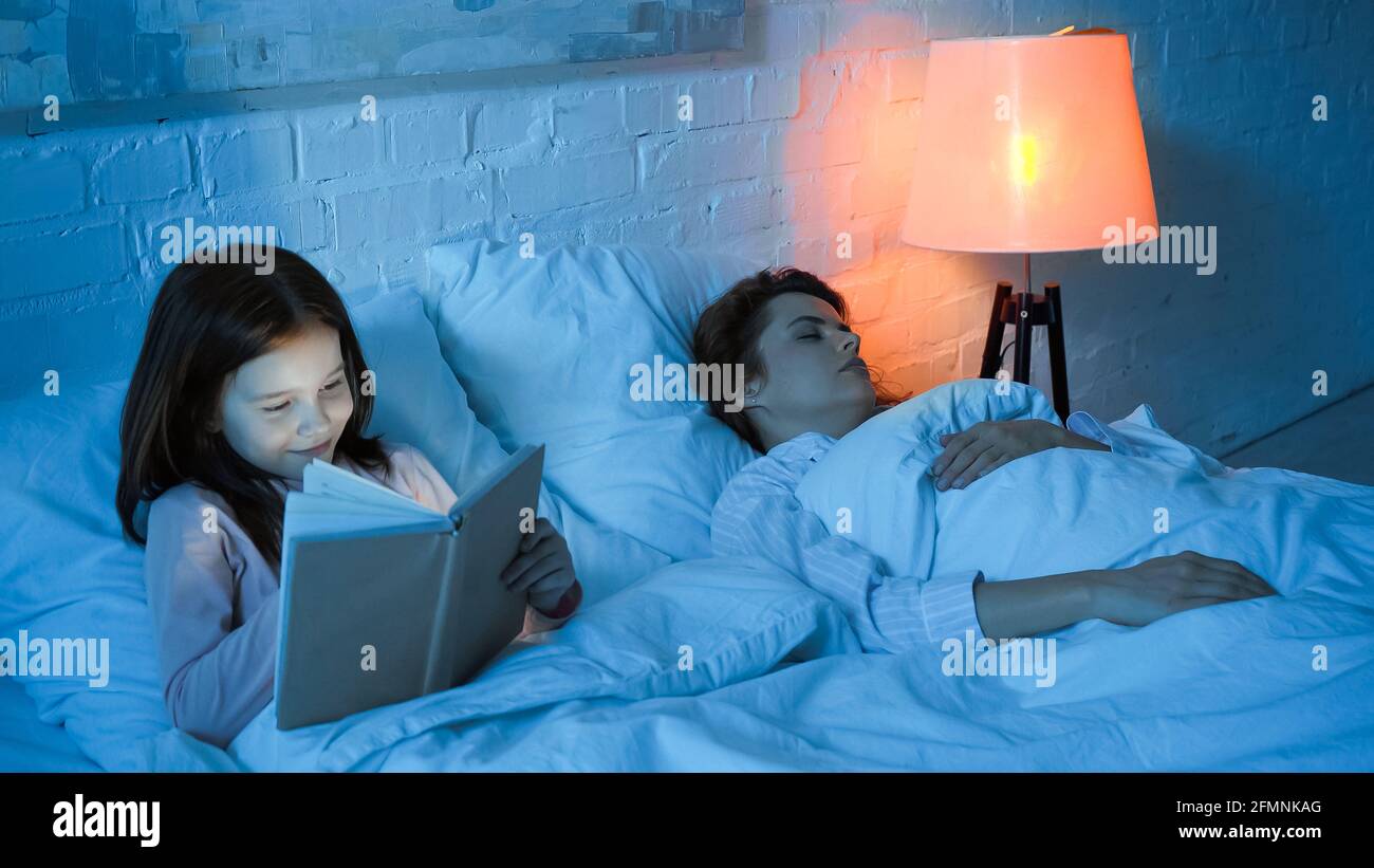 Smiling girl reading book while mother sleeping on bed Stock Photo - Alamy