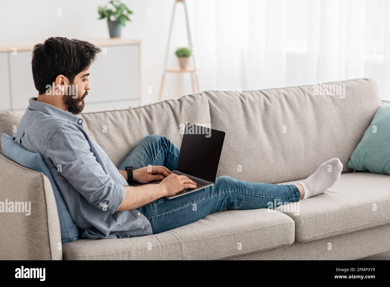 Relaxed arab man using laptop with blank screen, resting on couch in ...