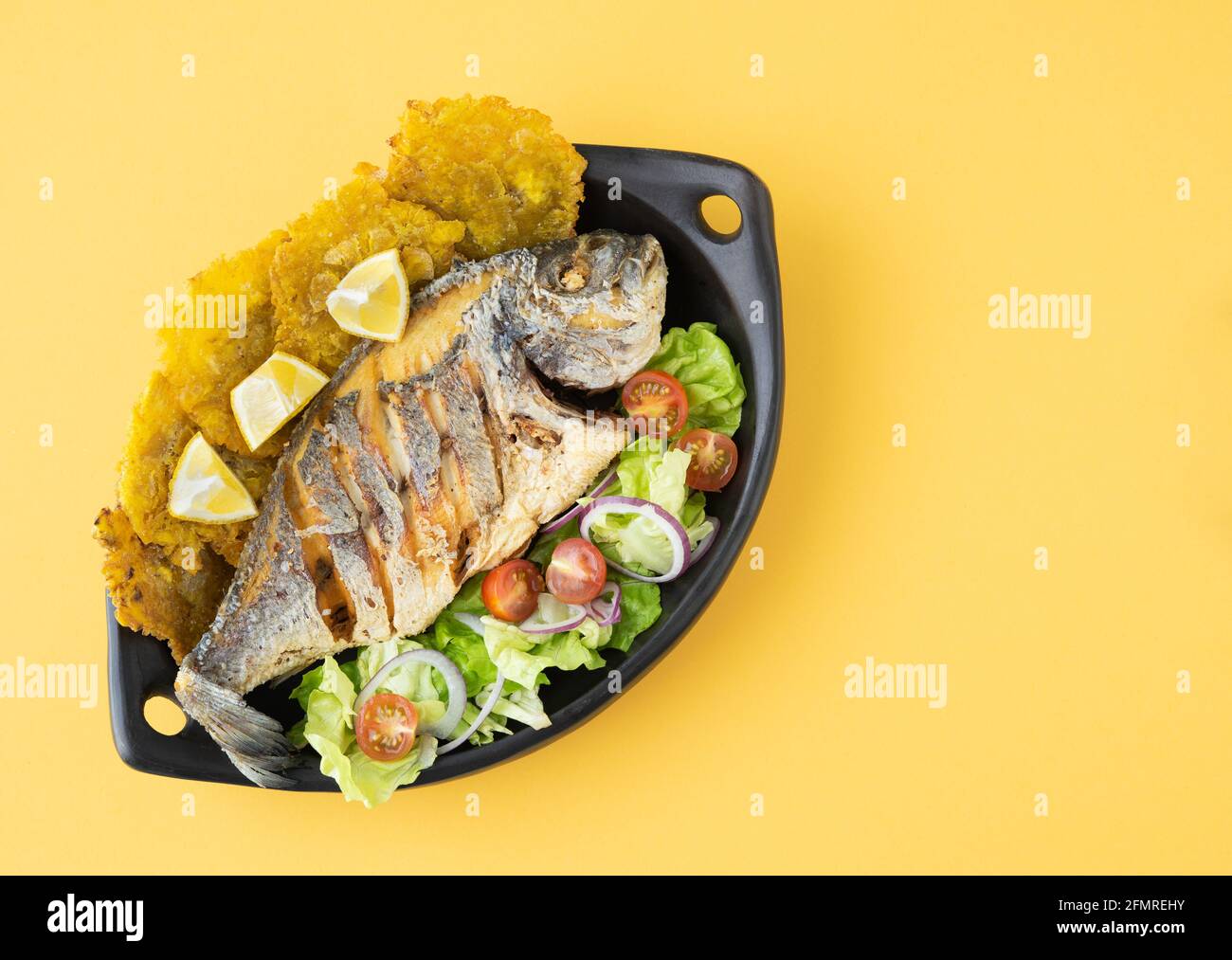 Fried fish with salad and patacones on black plate on yellow background ...
