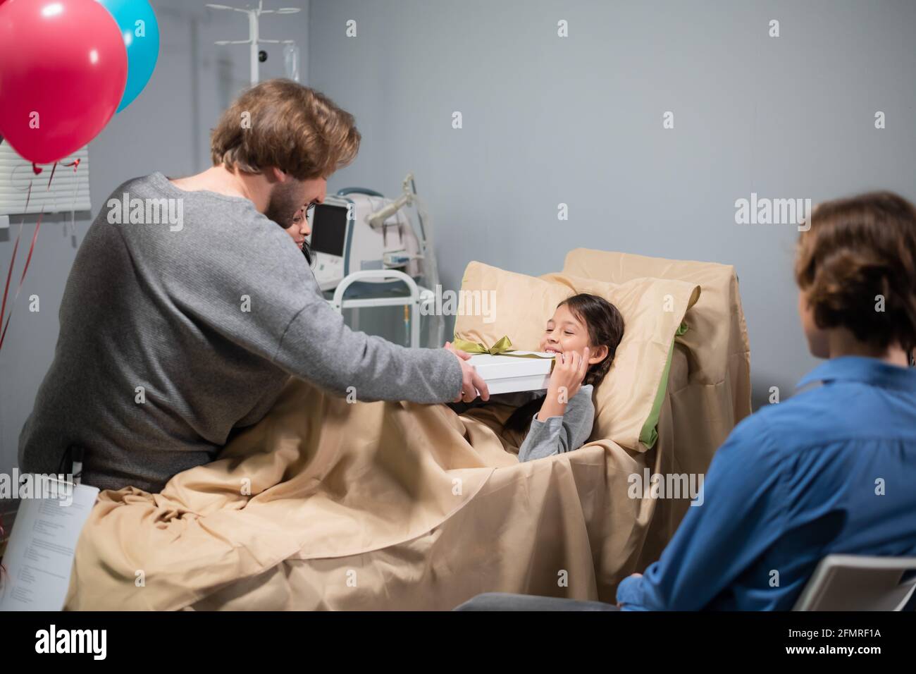 An older brother giving a little girl a present, she is lying in bed ...