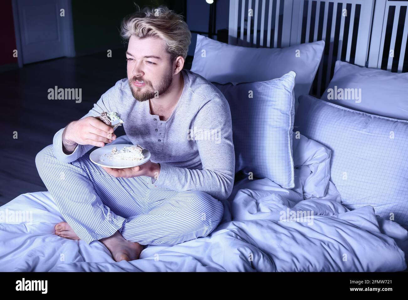 Young man eating cake while watching TV at night. Concept of addiction ...