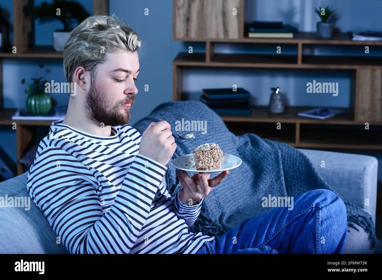 Young man eating cake while watching TV at night. Concept of addiction ...