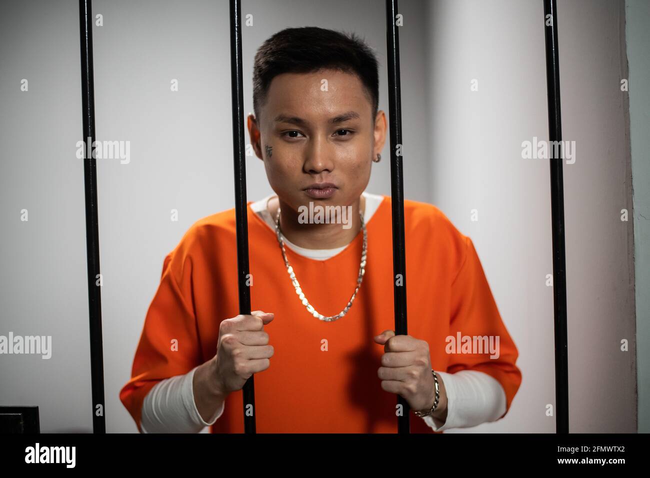 A young asian criminal poses for a photo against the bars in his cell ...
