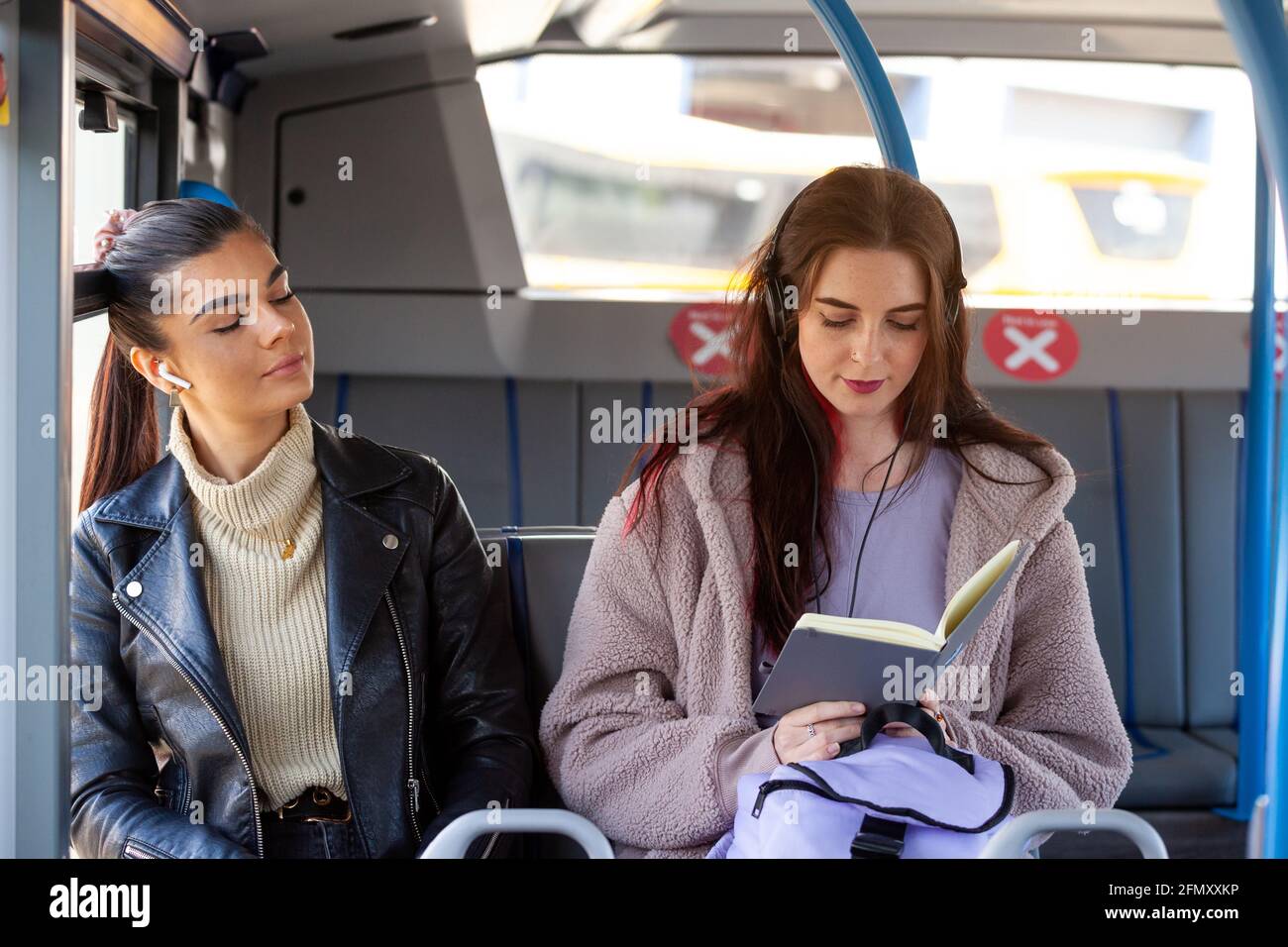 two women travelling on a bus both listening to music and one also ...