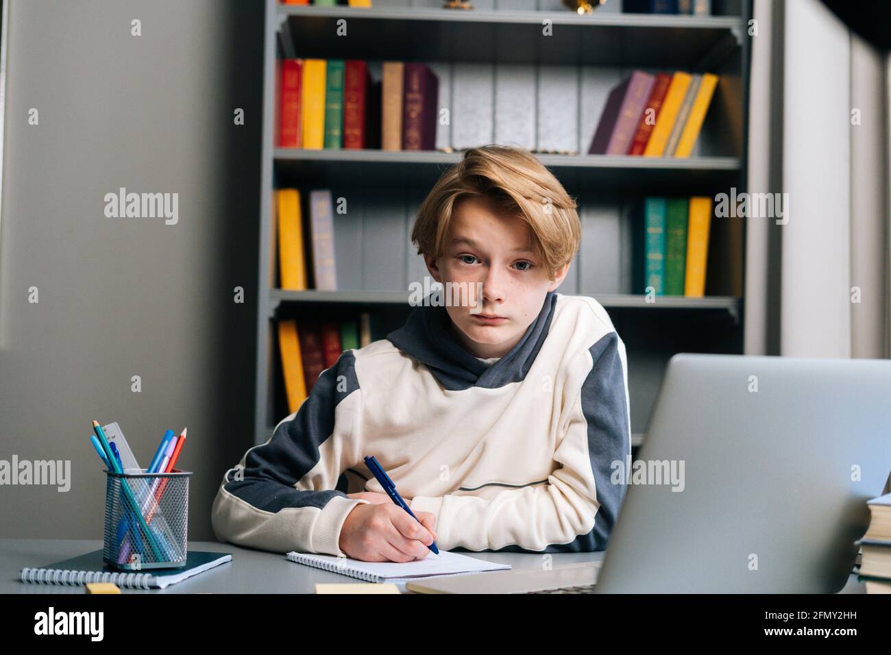 Portrait of pupil boy making notes in copybook with pen sitting at desk ...