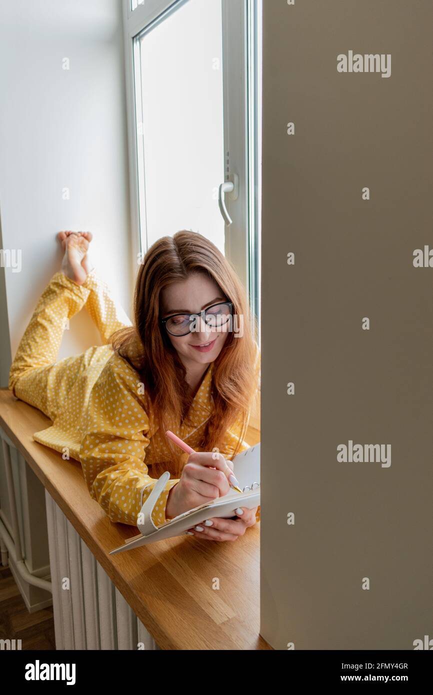 Young woman in comfortable yellow pajamas is working, lying down on the ...