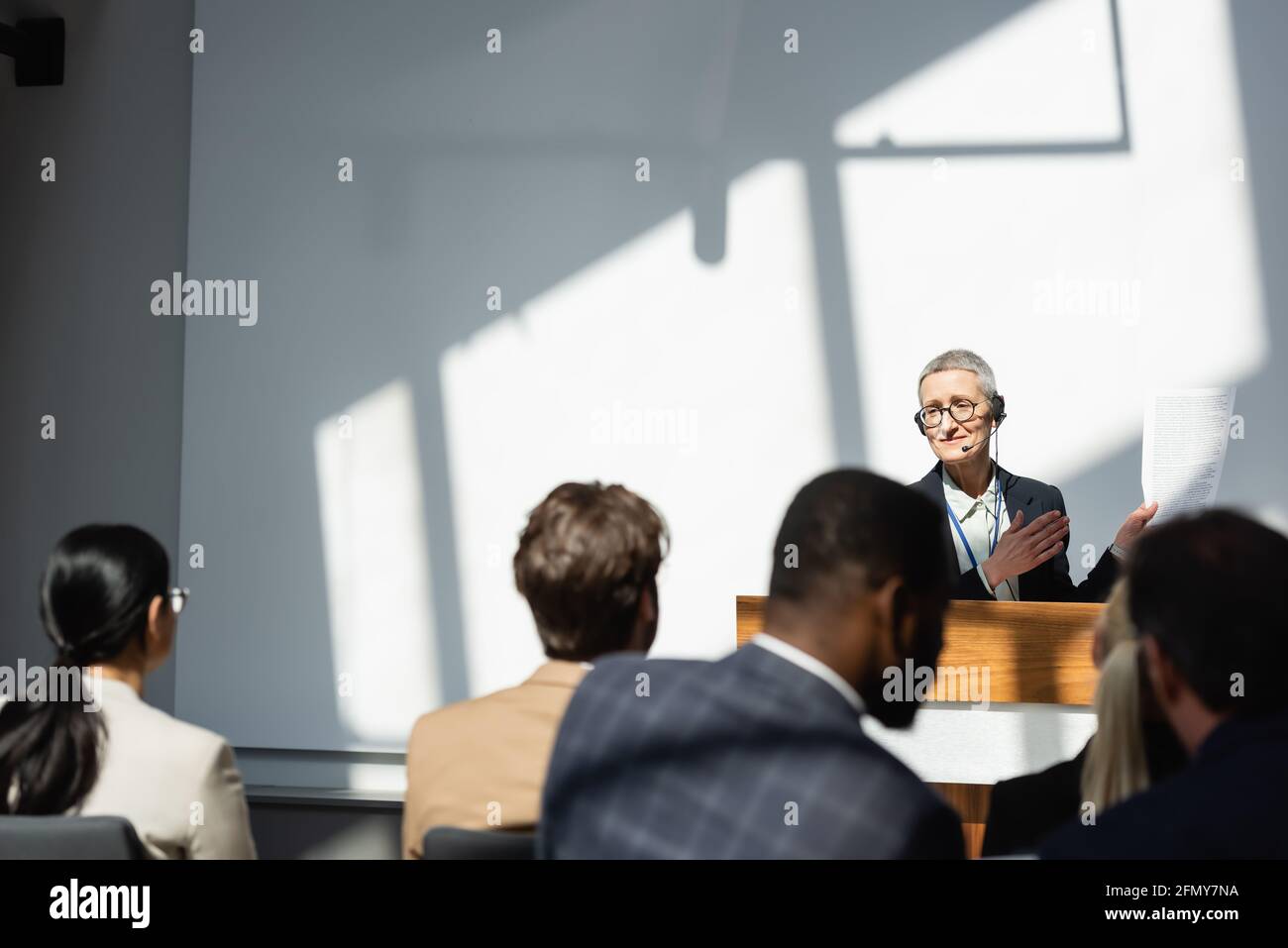 smiling lecturer pointing at document near blurred business people ...