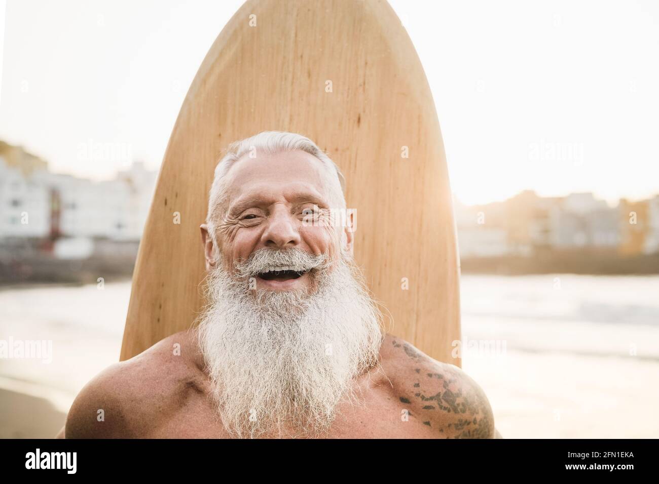 Tattooed senior surfer holding vintage surf board on the beach at ...