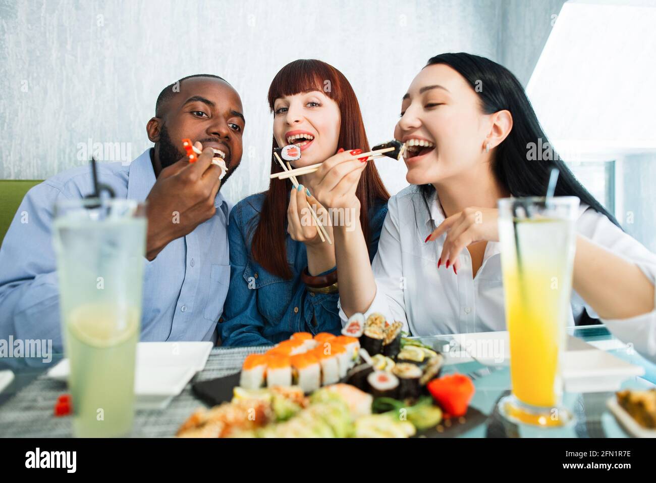 Young jolly multiracial friends eating sushi using chopsticks, looking ...