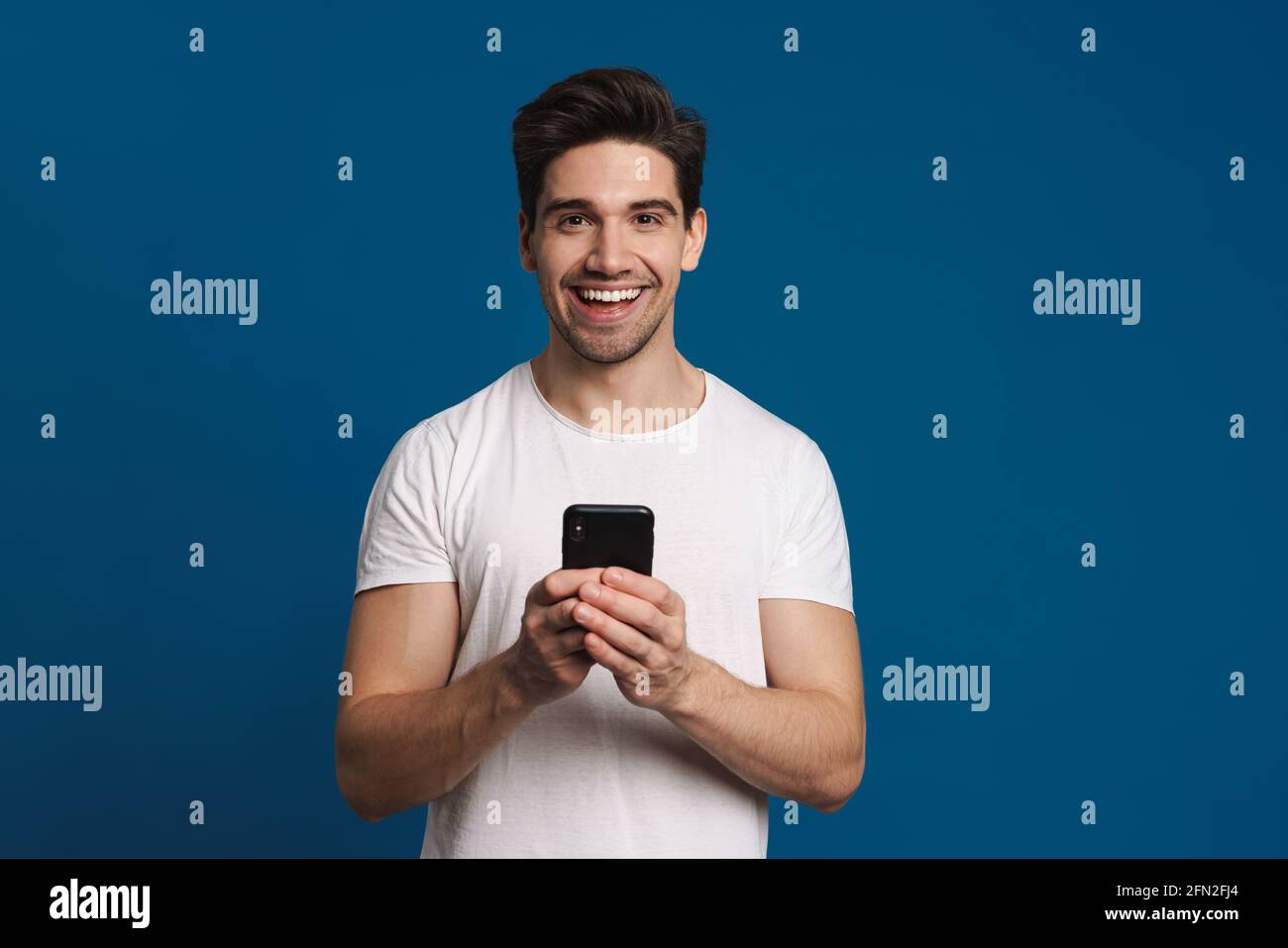 Happy unshaven guy smiling while using mobile phone isolated over blue ...