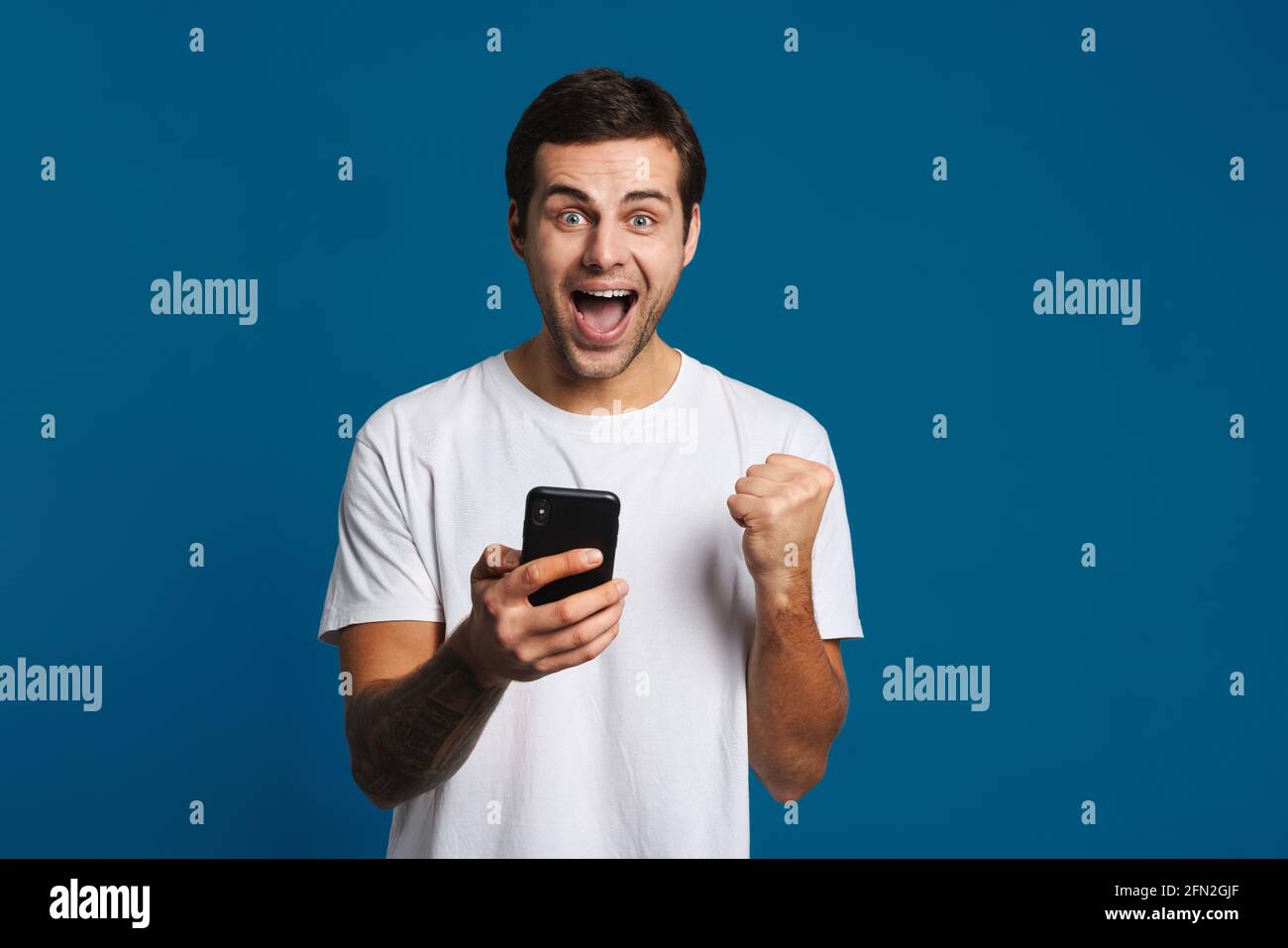 Delighted guy making winner gesture while using mobile phone isolated ...