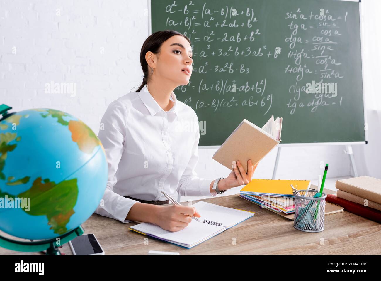 Teacher holding book and writing on notebook near globe in classroom ...