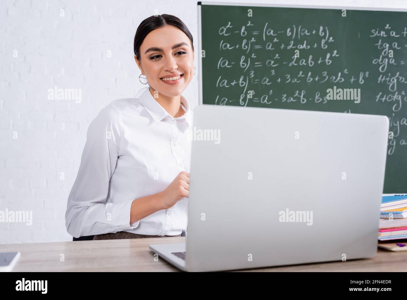 Positive teacher looking at blurred laptop in classroom Stock Photo - Alamy