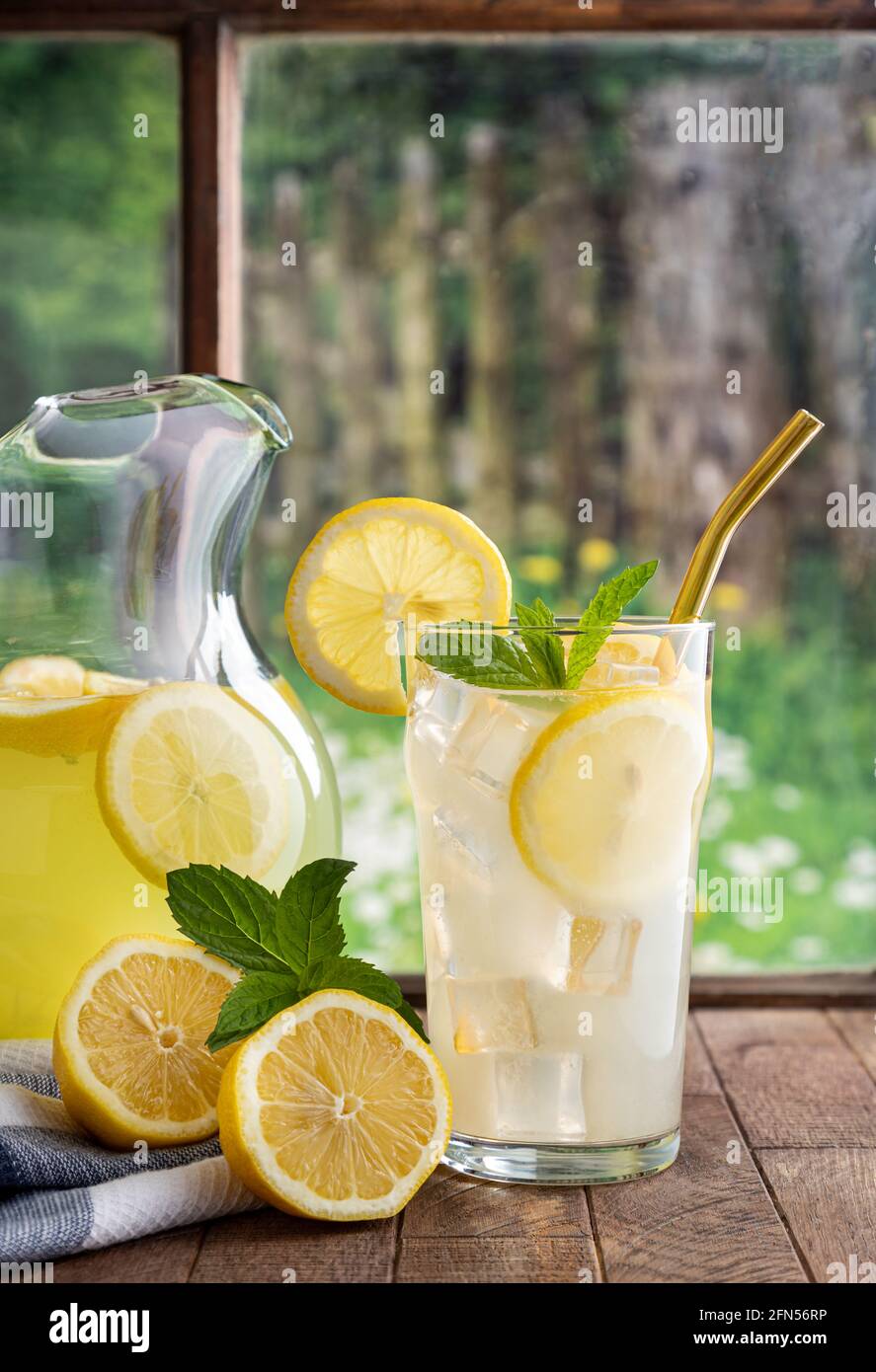 Glass of lemonade and pitcher with lemons on a wooden table by a window ...