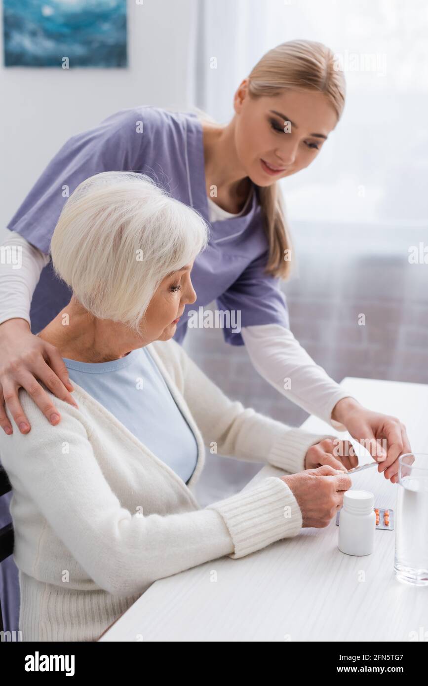 social worker touching shoulder of aged woman pointing at pills near ...
