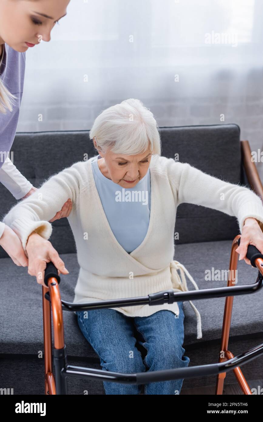 social worker helping elderly woman getting up from sofa near medical ...