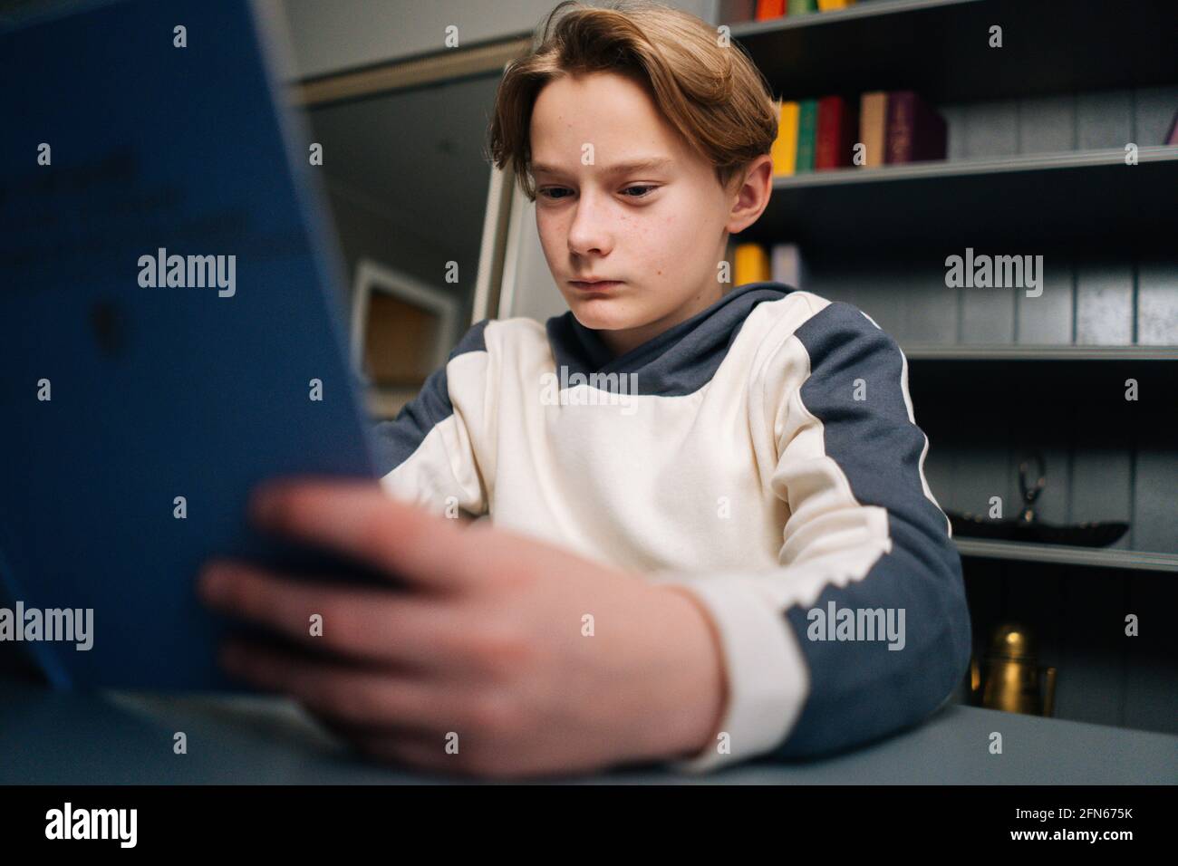 Close-up of focused serious pupil boy reading paper book sitting alone ...