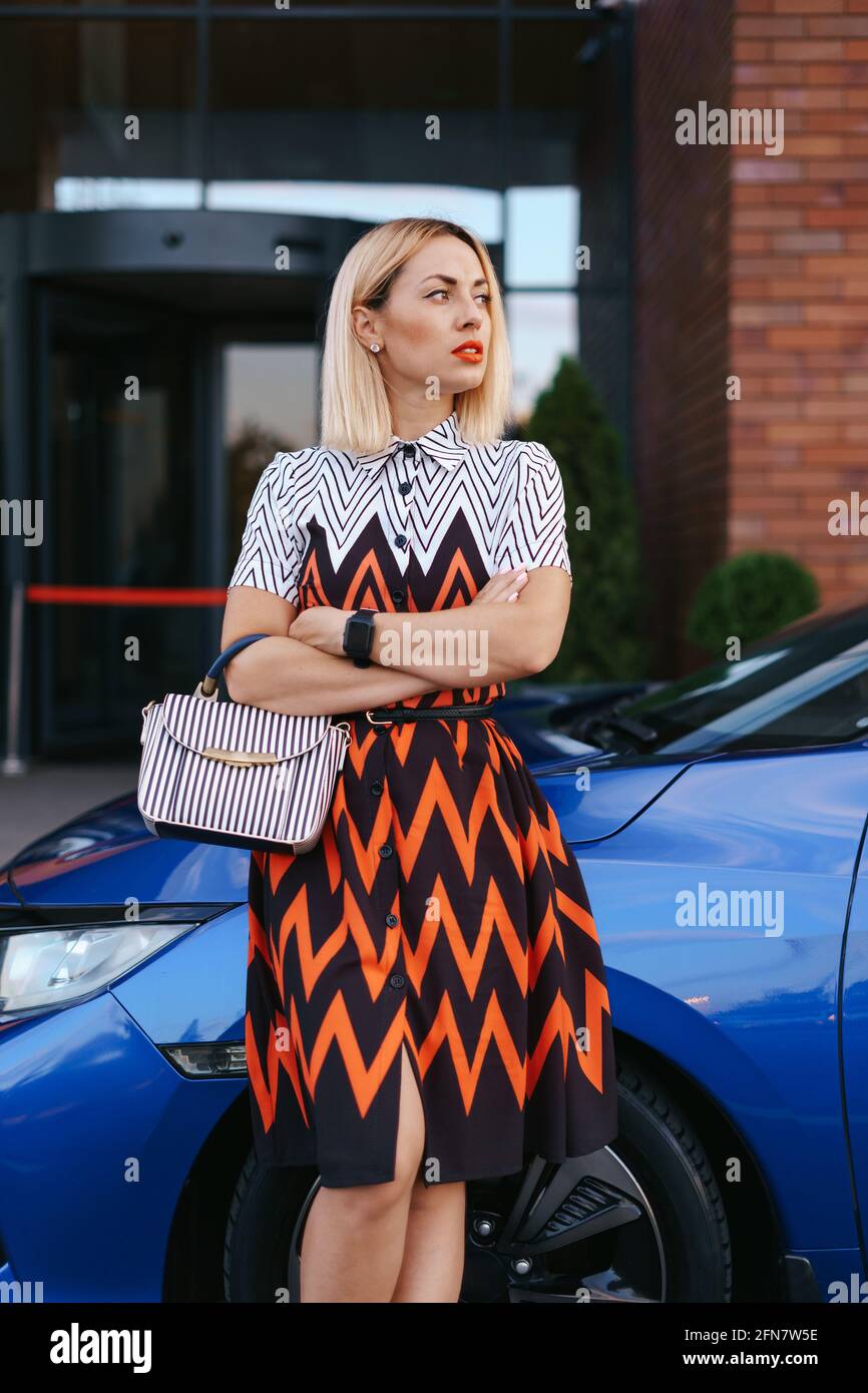 Stunning young woman waring dress posing in front of her car outdoors ...