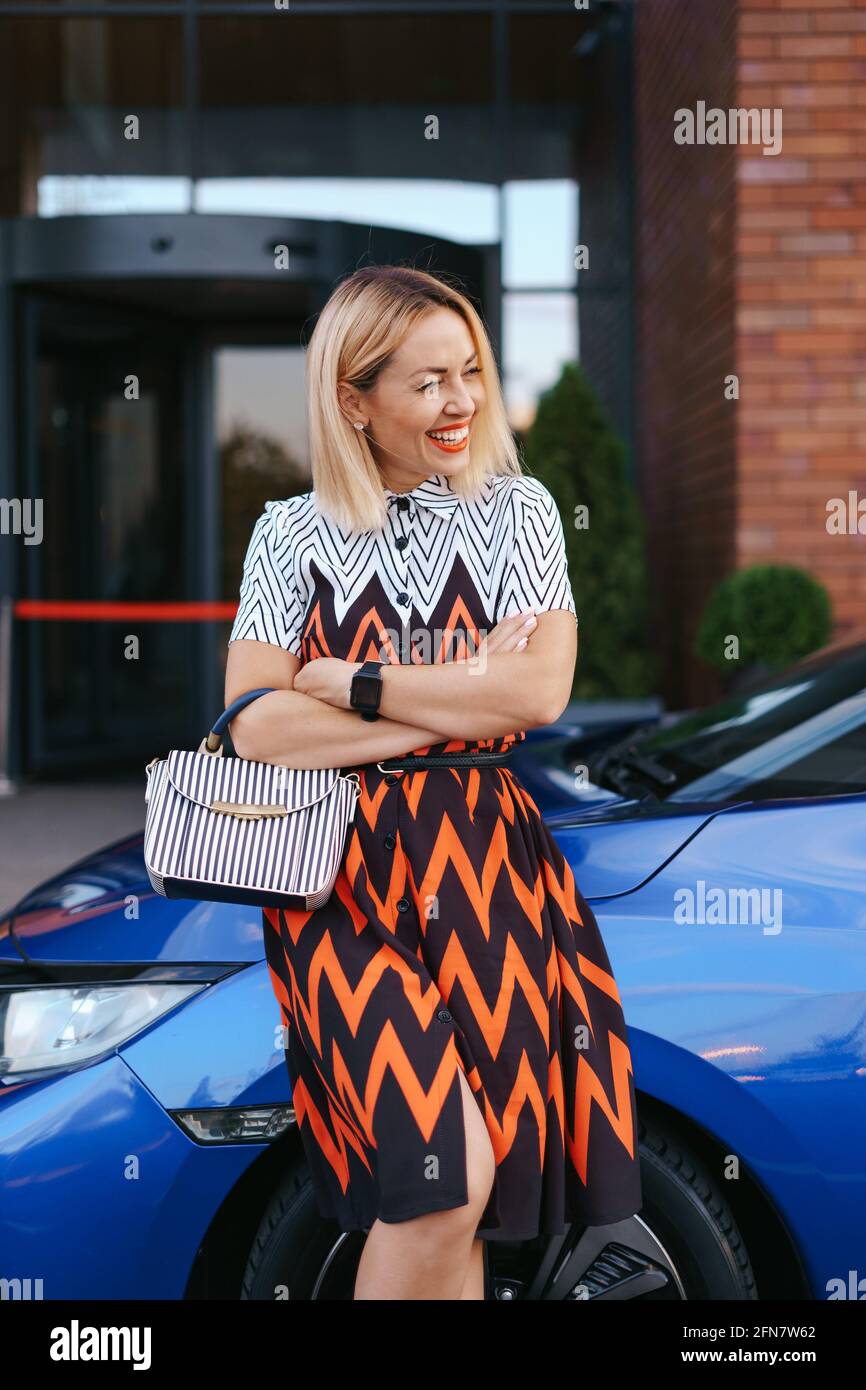 Stunning young woman waring dress posing in front of her car outdoors ...