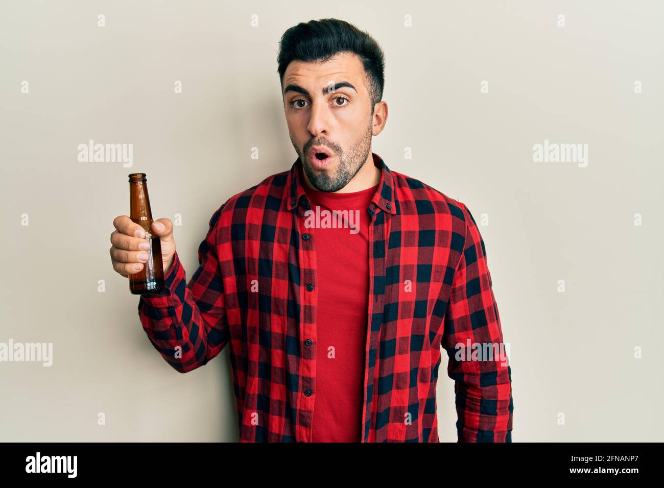 Young hispanic man drinking a bottle of beer scared and amazed with ...